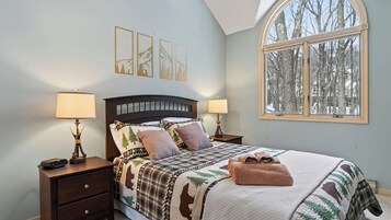 Quarto com uma cama grande, duas mesas de cabeceira, uma janela com vista para árvores e três quadros pendurados na parede.