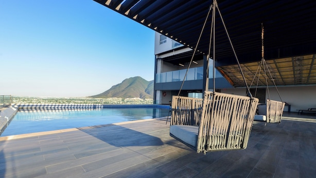 Uma área de piscina no terraço com vista para uma montanha, uma cadeira suspensa e uma estrutura de prédio moderna.