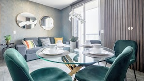 Uma sala de estar moderna com uma mesa de jantar de vidro, cadeiras de veludo verde, um sofá cinza e uma janela grande com persianas de madeira.