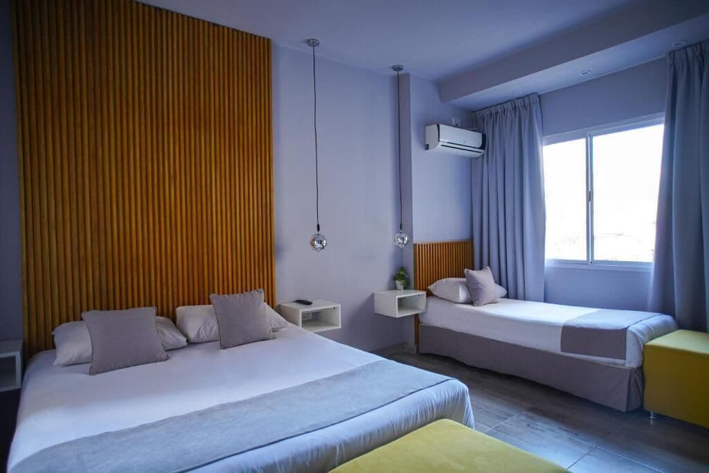 Quarto de hotel com duas camas, uma janela com cortinas, ar condicionado fixado na parede e uma pequena mesa de cabeceira com abajur.