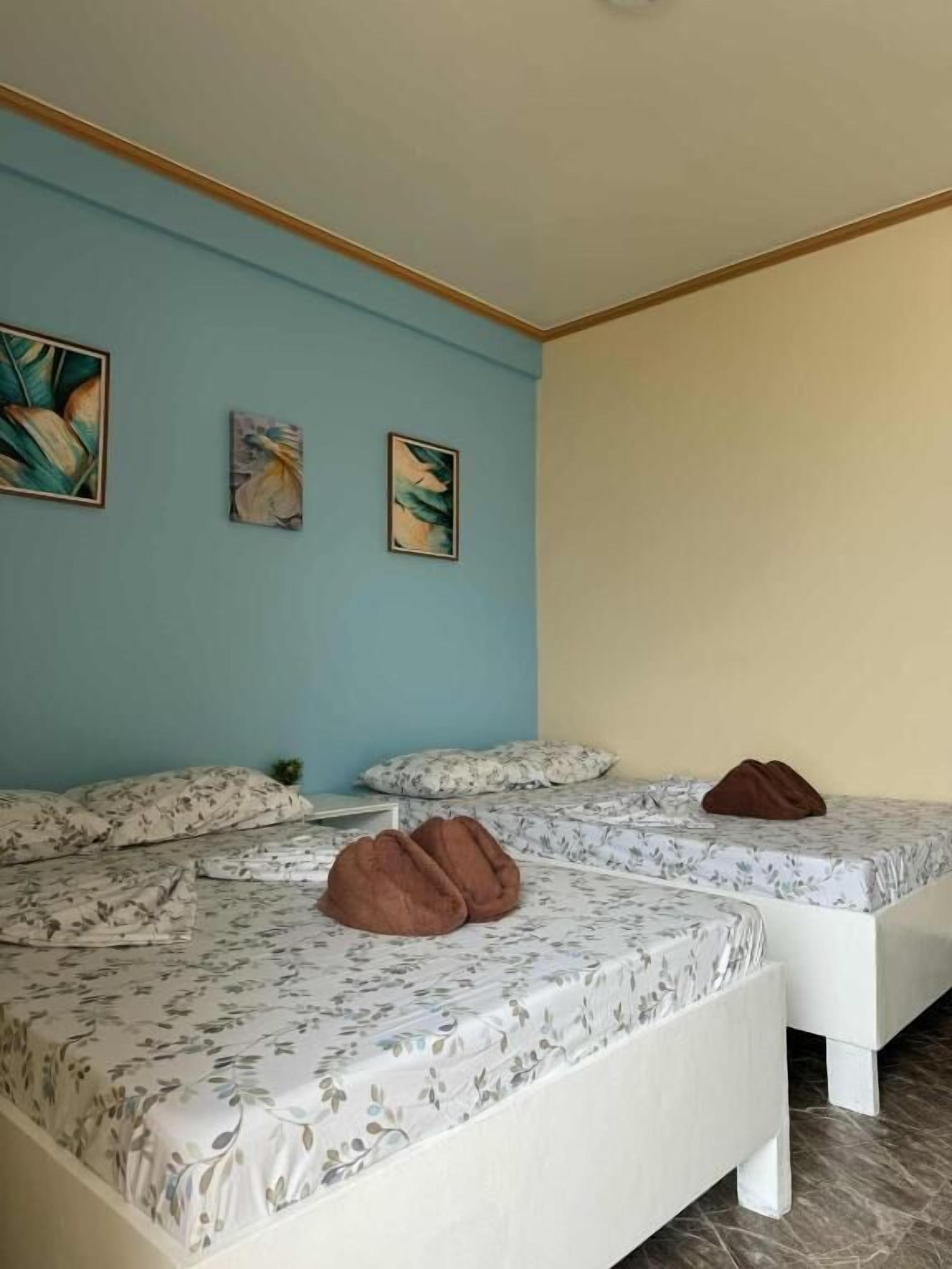 Quarto com duas camas, cada uma com roupa de cama estampada com flores e almofadas castanhas. Três quadros pendurados na parede, e o quarto tem uma paleta de cores azul claro e bege.