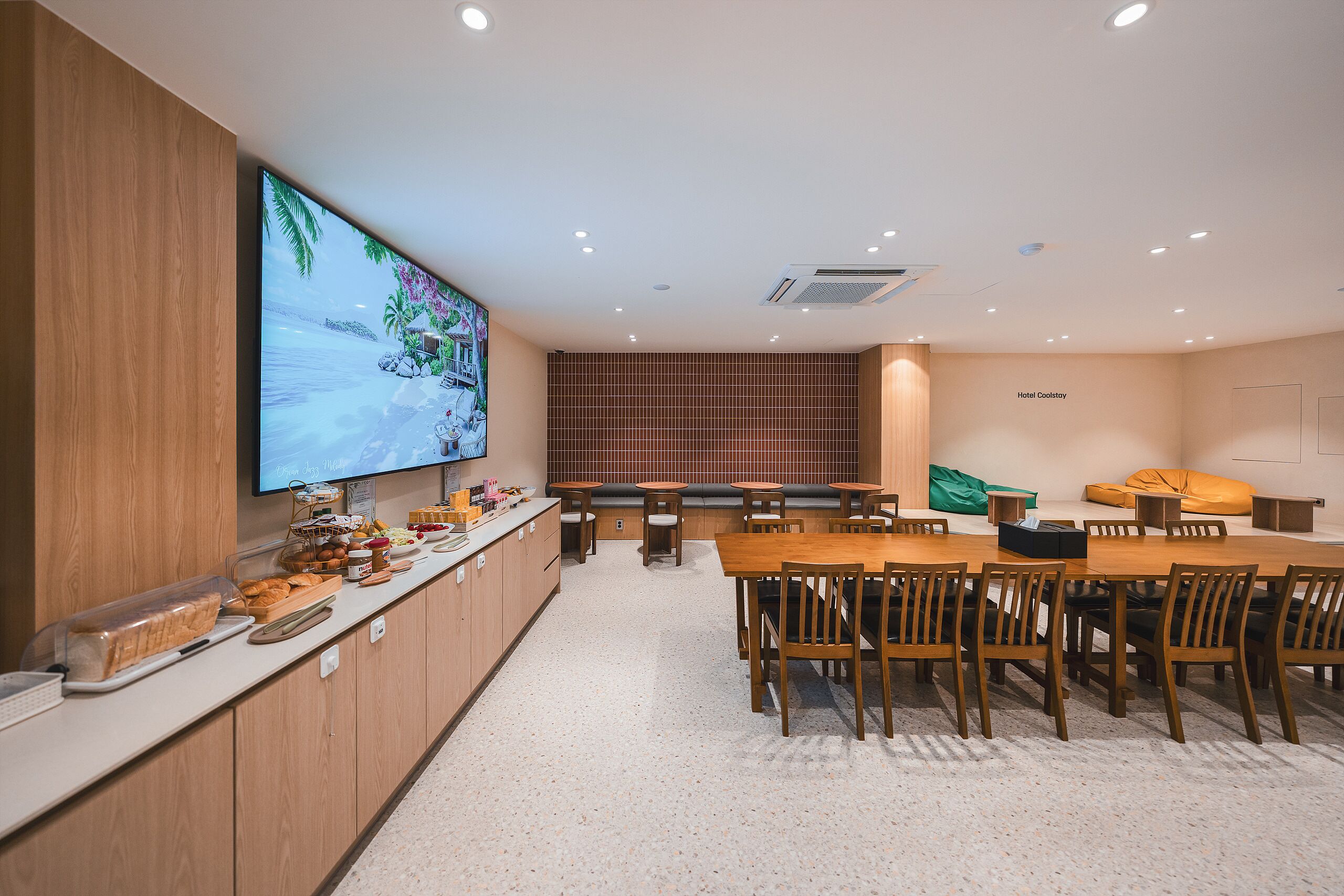 Quarto de hotel moderno com uma televisão de tela plana grande, um aparador de madeira, uma área de jantar com uma mesa longa e cadeiras, e uma parede com padrão quadriculado.