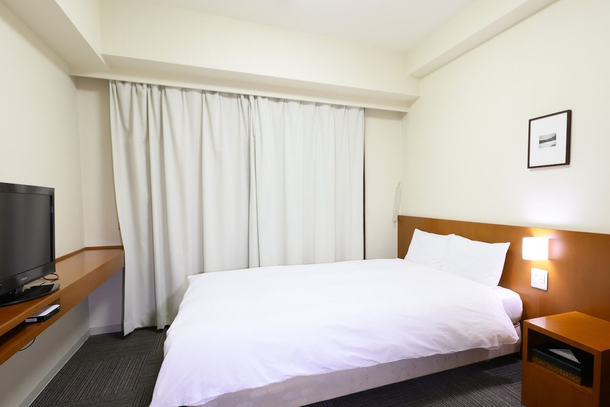Quarto de hotel com cama, televisão, mesa de cabeceira, luminária e um quadro na parede.