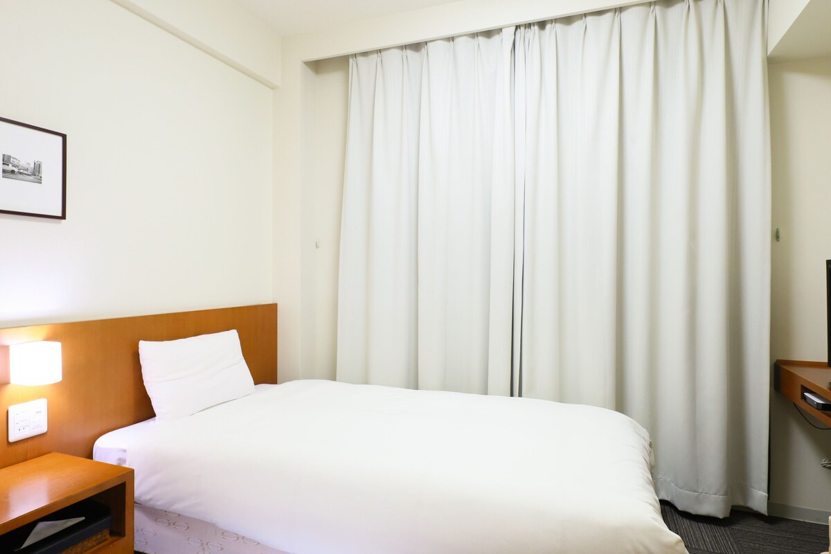 Quarto de hotel com cama, mesa de cabeceira, televisão e cortina.