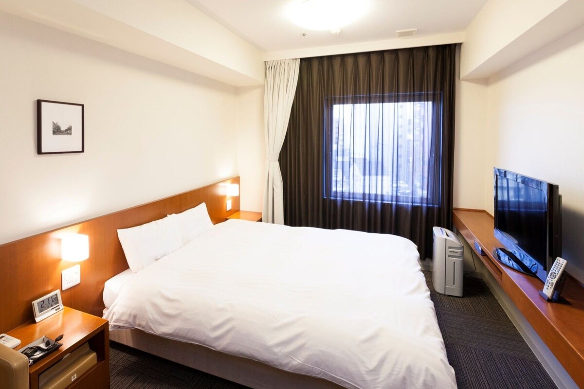 Quarto de hotel com cama, luminárias de cabeceira, televisão, escrivaninha com computador e janela com cortinas.