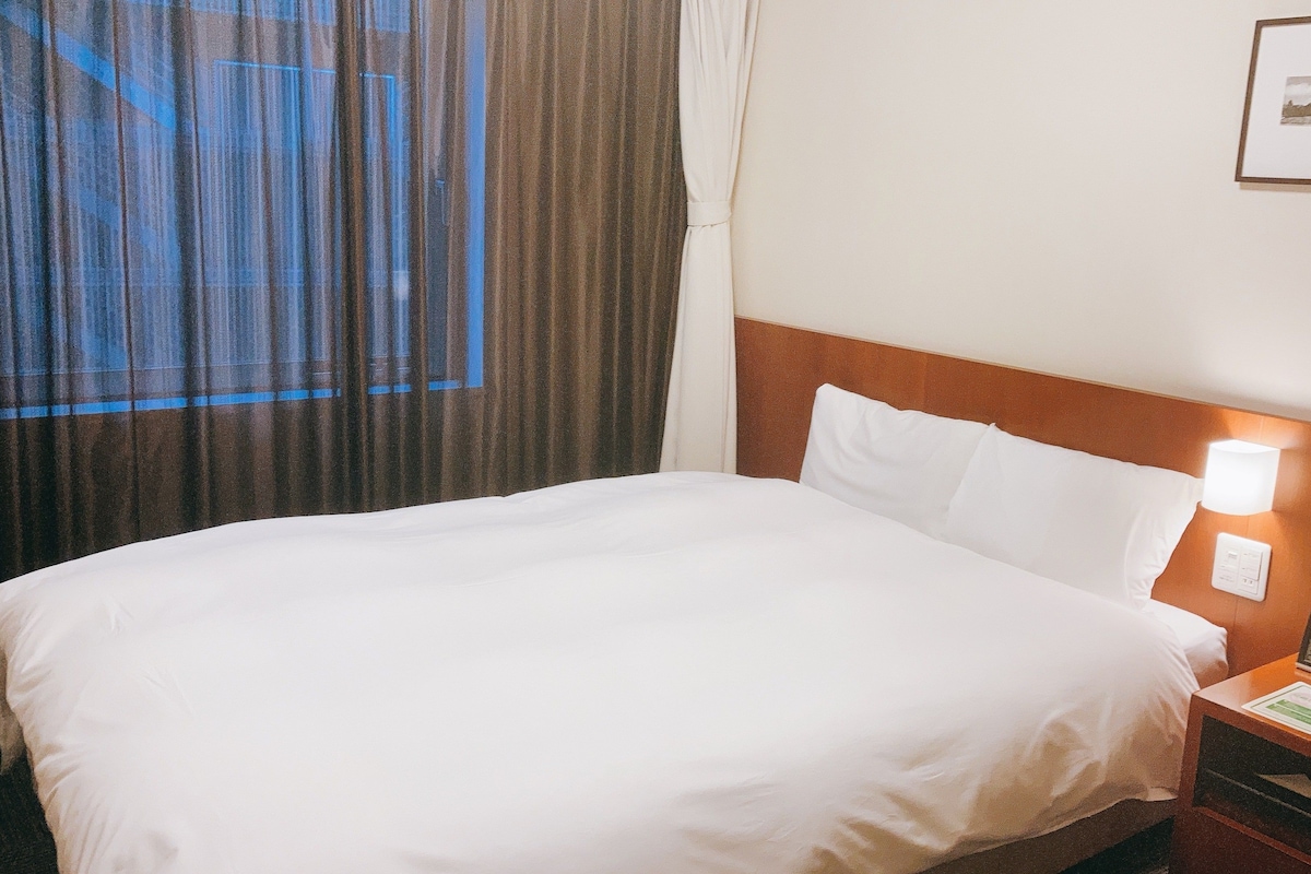 Quarto de hotel com cama, mesa de cabeceira com relógio e quadro na parede.