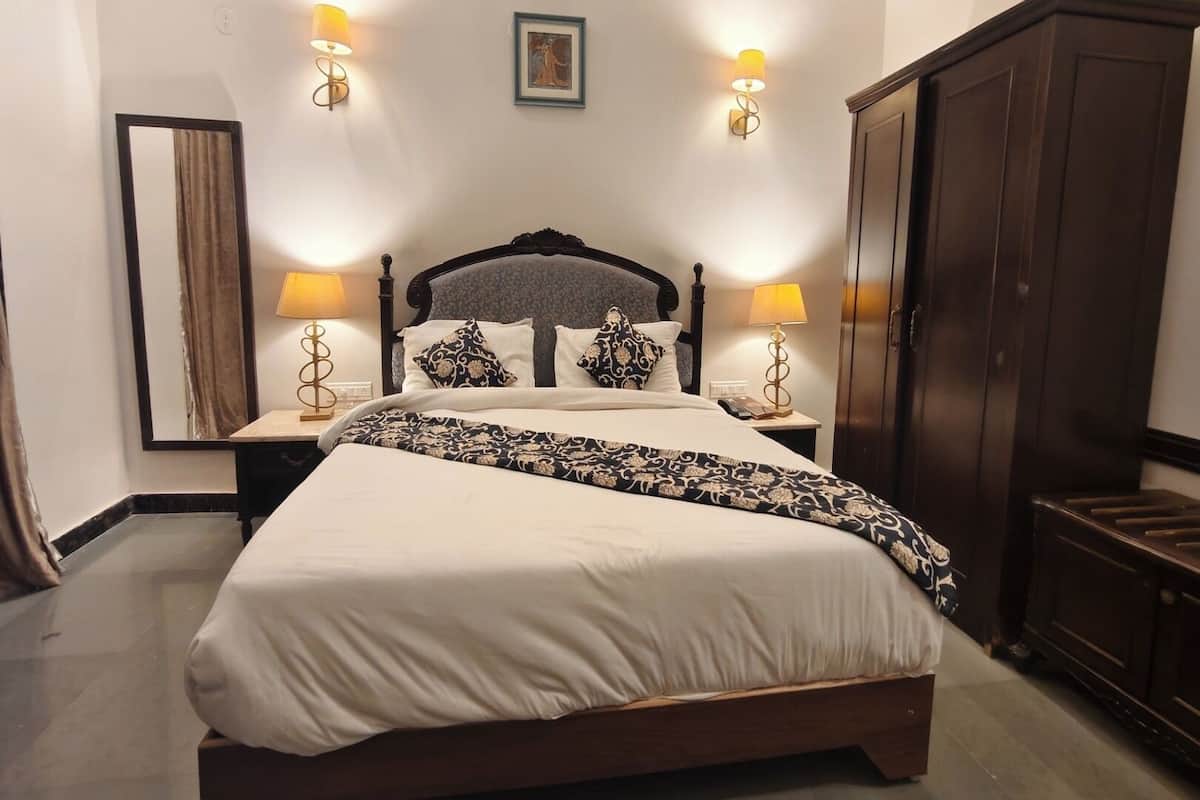 Cama bem arrumada com colcha estampada, duas luminárias de cabeceira, um quadro emoldurado acima da cama e um guarda-roupa de madeira escura à direita.
