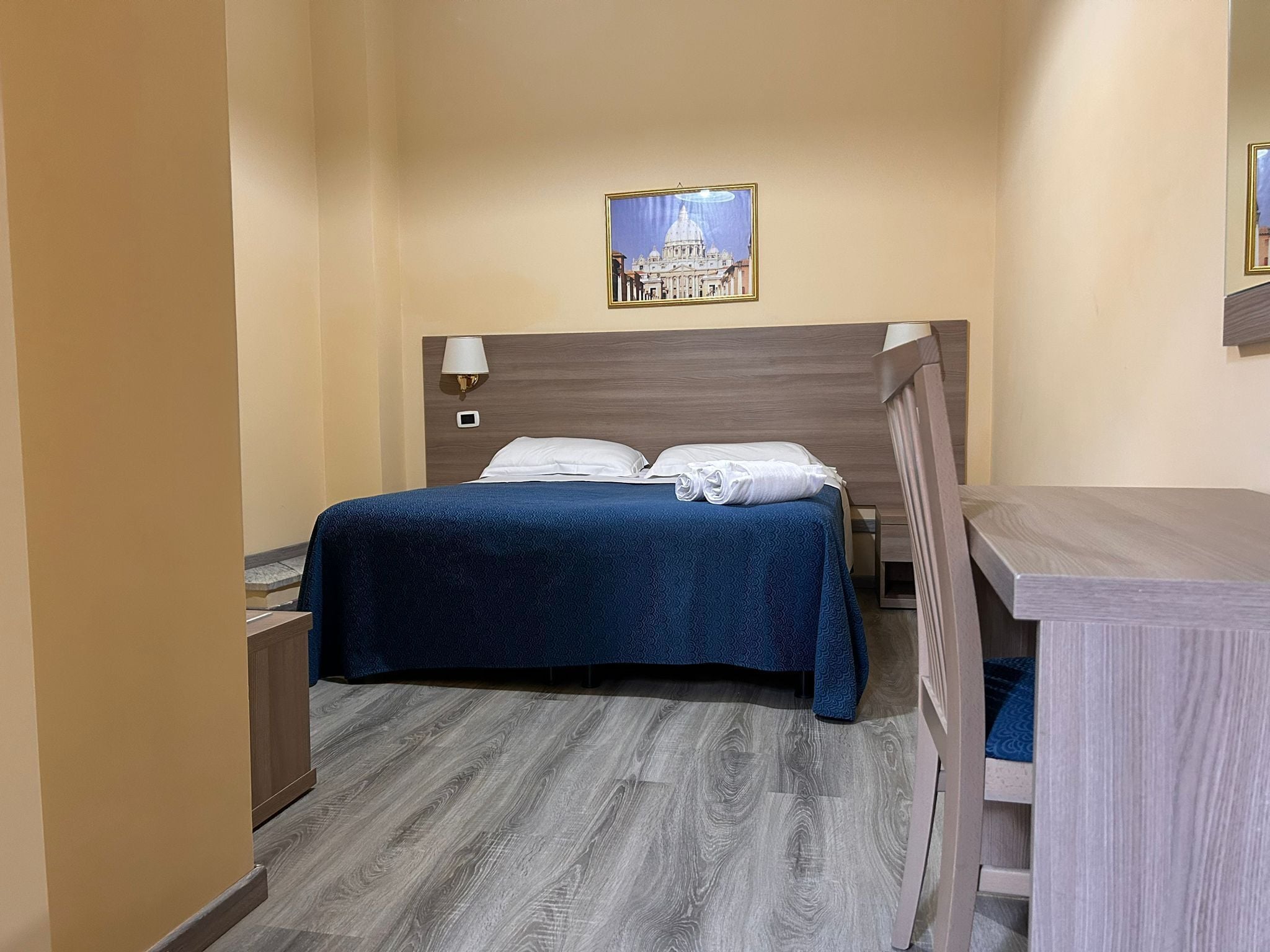 Quarto de hotel com uma cama, uma escrivaninha, uma mesa de cabeceira, uma luminária na parede e um quadro com a imagem de um edifício.