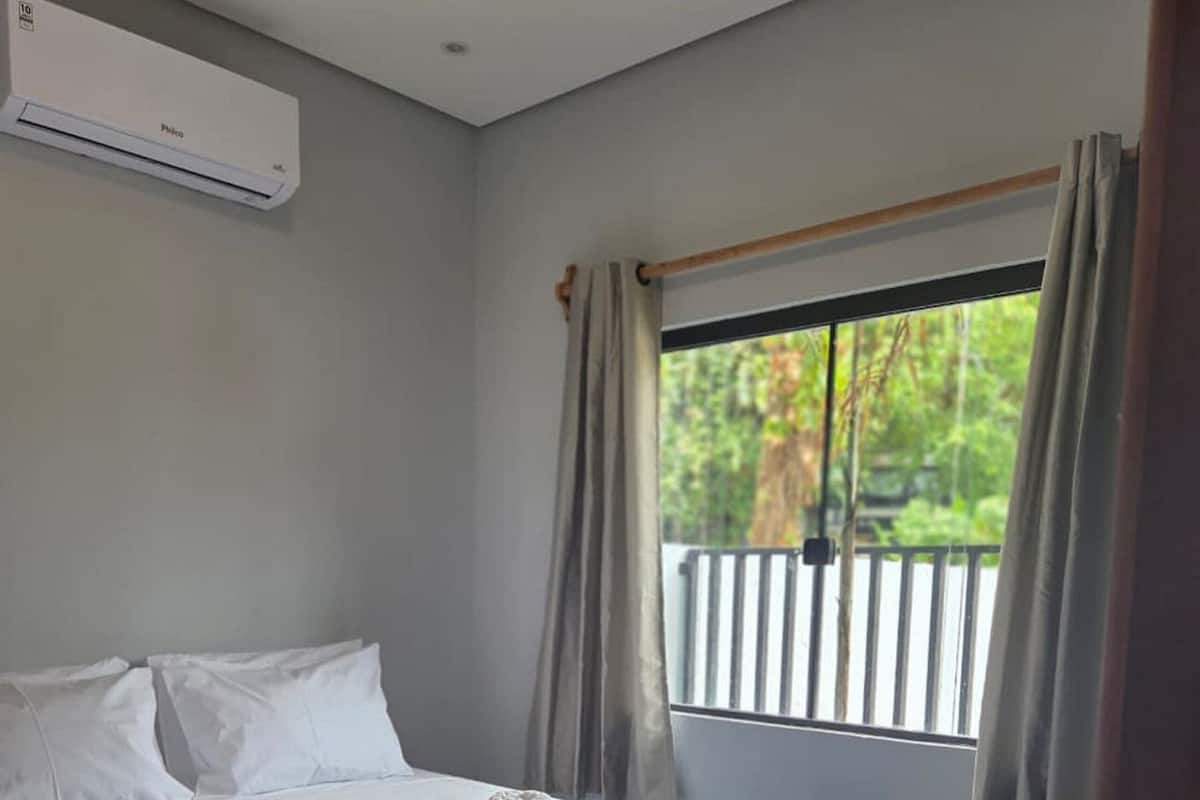 Quarto de hotel com cama, janela com cortinas, ar condicionado branco e vista para o verde lá fora.