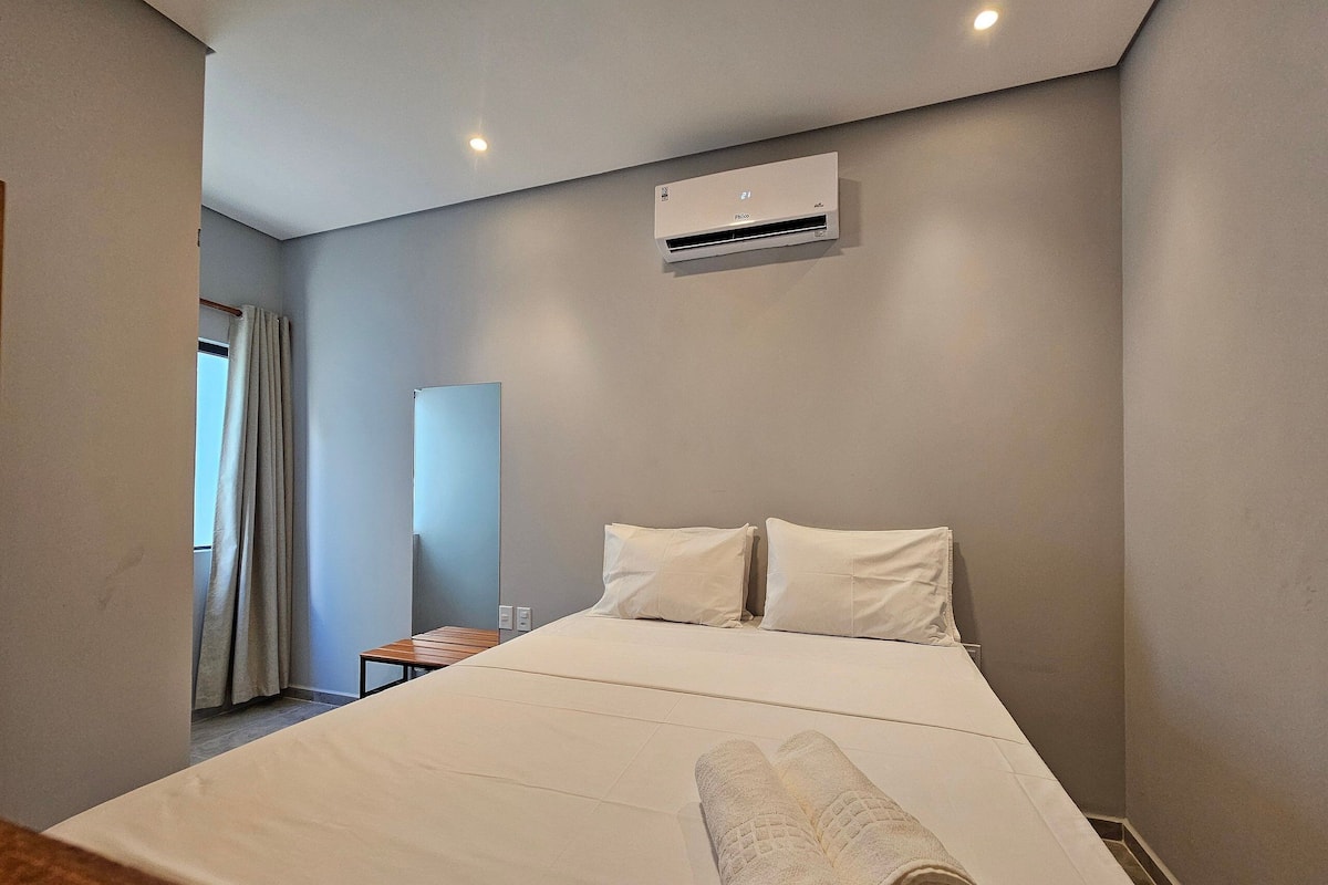 Quarto de hotel com cama, ar condicionado na parede e janela com cortinas.