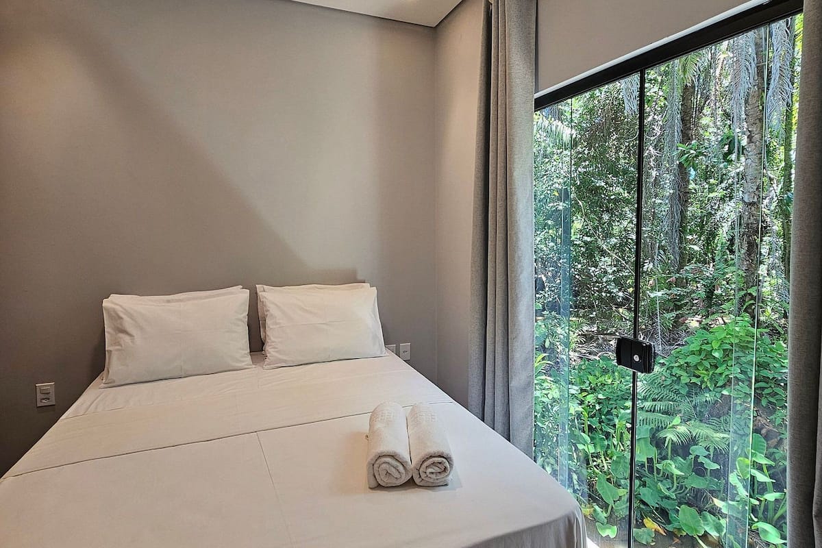 Quarto de hotel moderno com uma cama grande, vista para uma floresta através de janelas do chão ao teto e uma luminária no teto.