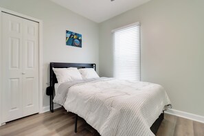Quarto com cama, porta branca, janela com persianas e uma obra de arte na parede.