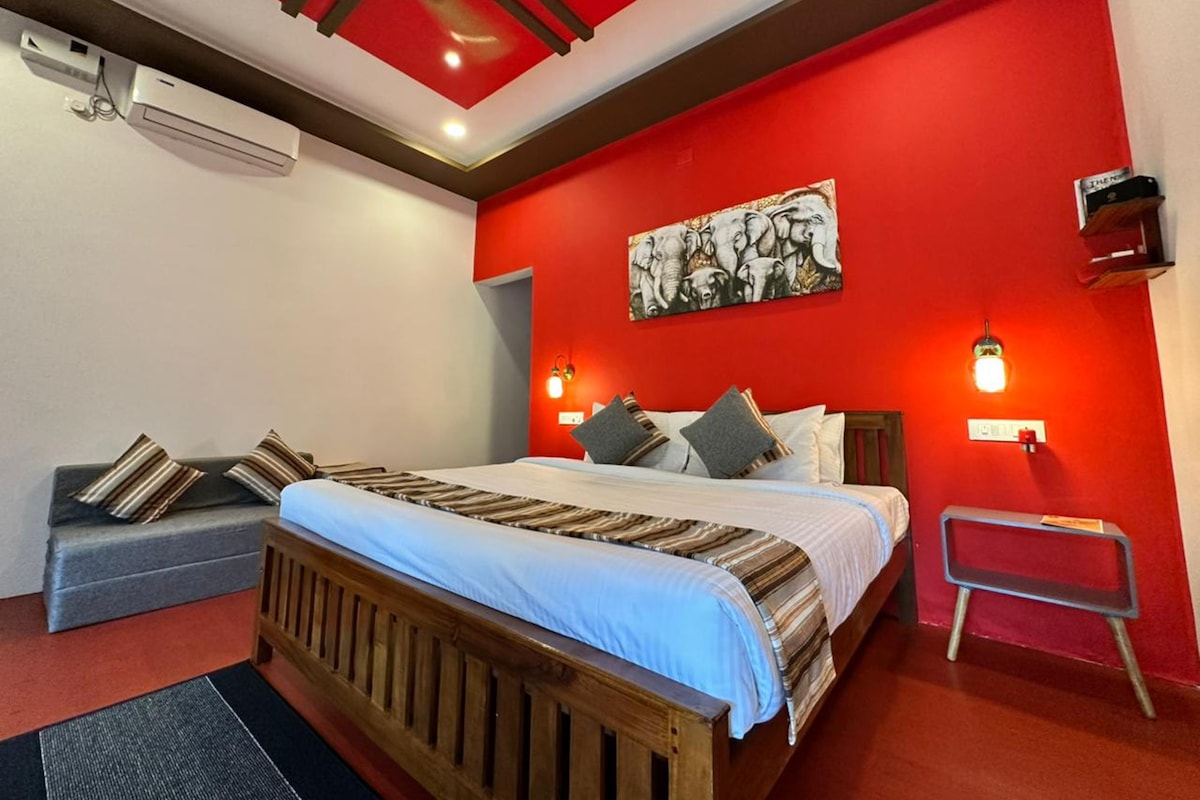 Quarto de hotel com cama de madeira, paredes vermelhas, ventoinha no teto e pintura de elefantes.