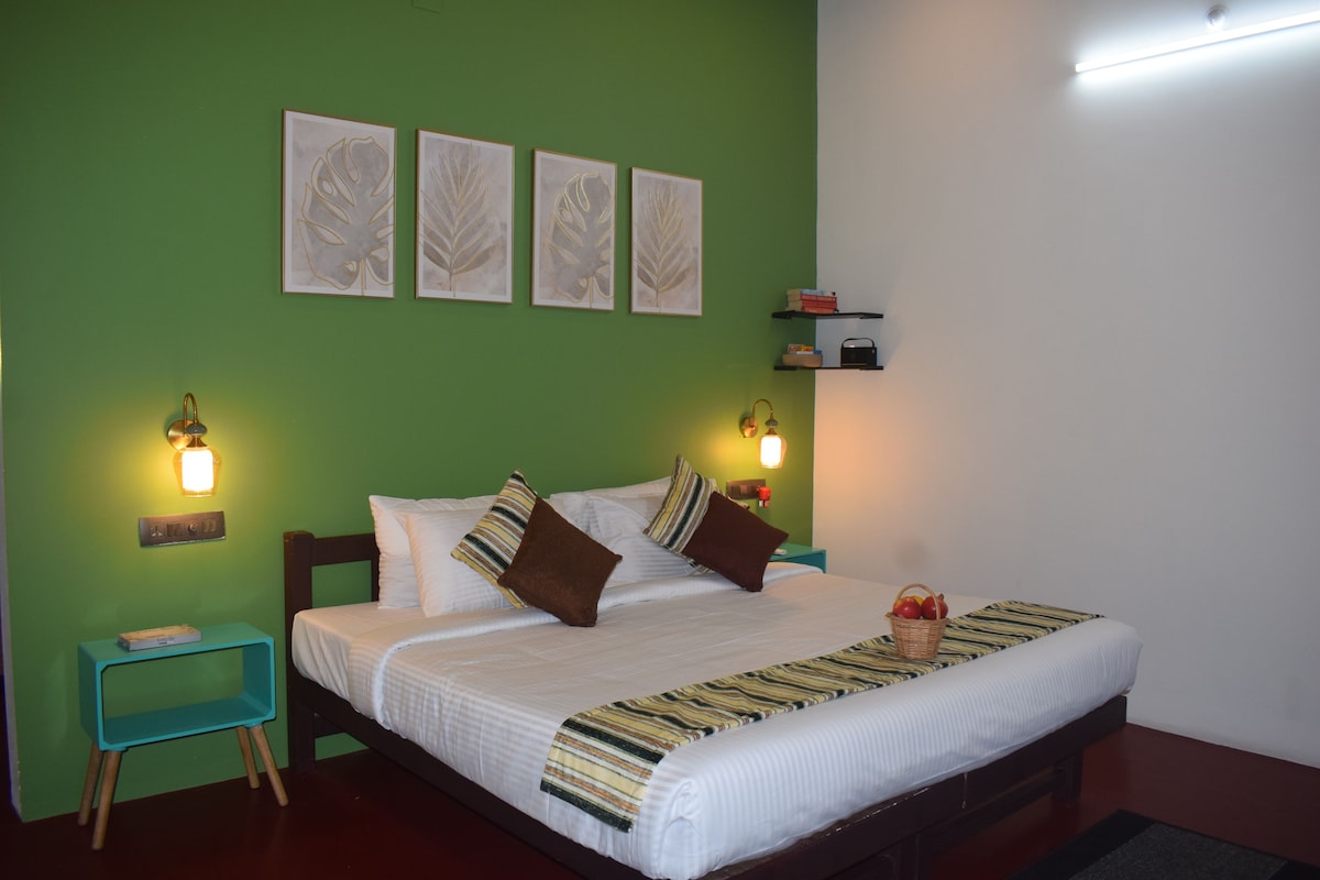 Cama bem arrumada com travesseiros e manta listrada, uma pequena mesa de cabeceira, prateleira na parede com livros e rádio, e três obras de arte emolduradas.