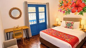 Quarto de hotel com uma cama, escrivaninha, cadeira, porta azul e um mural floral na parede.