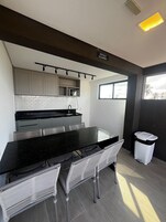 Uma cozinha moderna com bancada preta, armários brancos e micro-ondas. Há uma área de jantar com mesa preta e cadeiras brancas. Uma lixeira é visível perto da janela.
