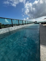 Piscina infinita no terraço, com guarda-corpo de vidro, proporcionando vista para a paisagem urbana e palmeiras.