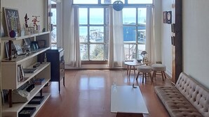 Uma sala de estar ampla com uma janela grande, um sofá branco, uma mesa de centro e uma estante.