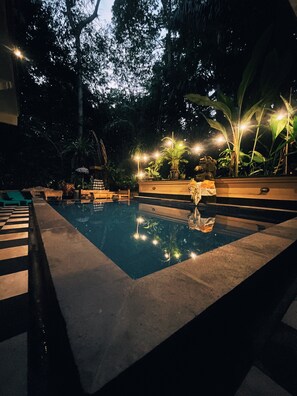 Área de piscina externa à noite, com borda de pedra, plantas em vasos e iluminação ambiente.