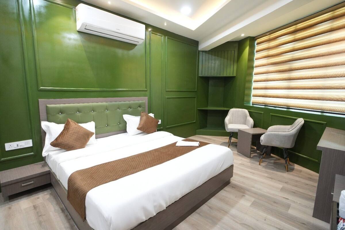 Quarto de hotel com uma cama grande, um espaço de estar com duas cadeiras, uma mesinha e uma janela com persianas.