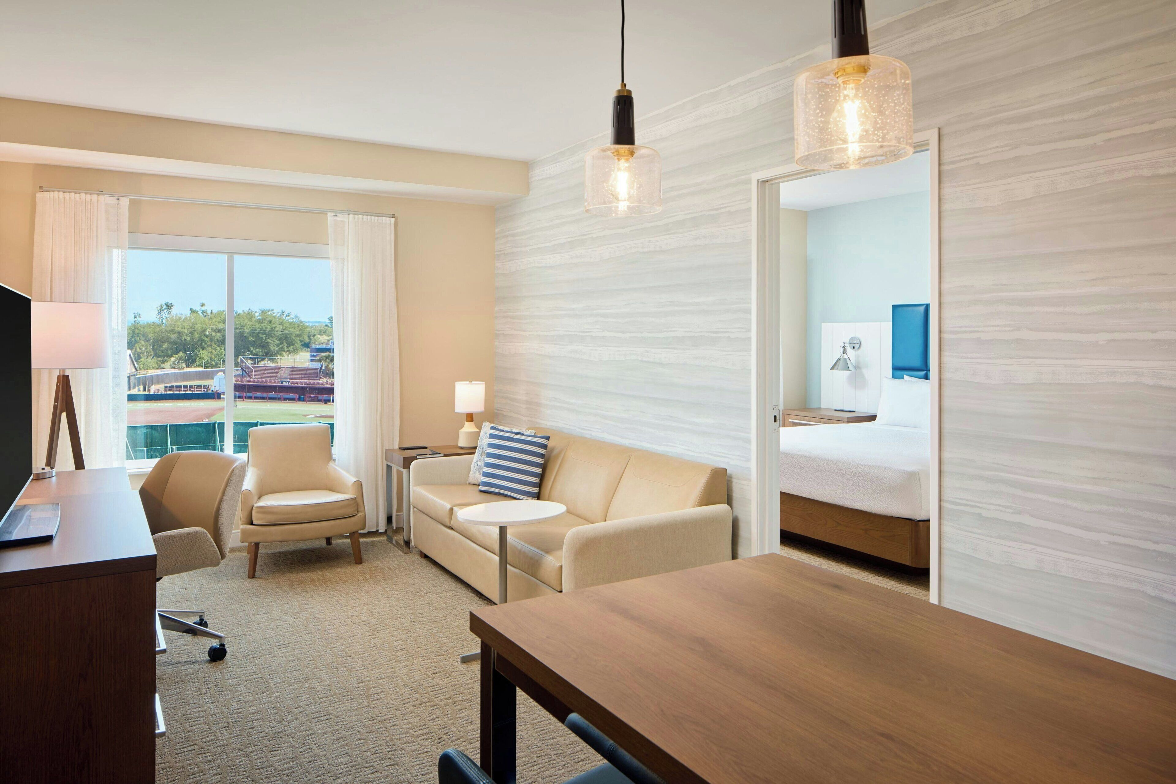 Quarto de hotel com escrivaninha, sofá, mesa pequena, cama e uma janela grande com cortinas.