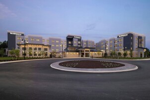 Hotel com vários andares, entrada central, caminho com paisagismo e céu claro.