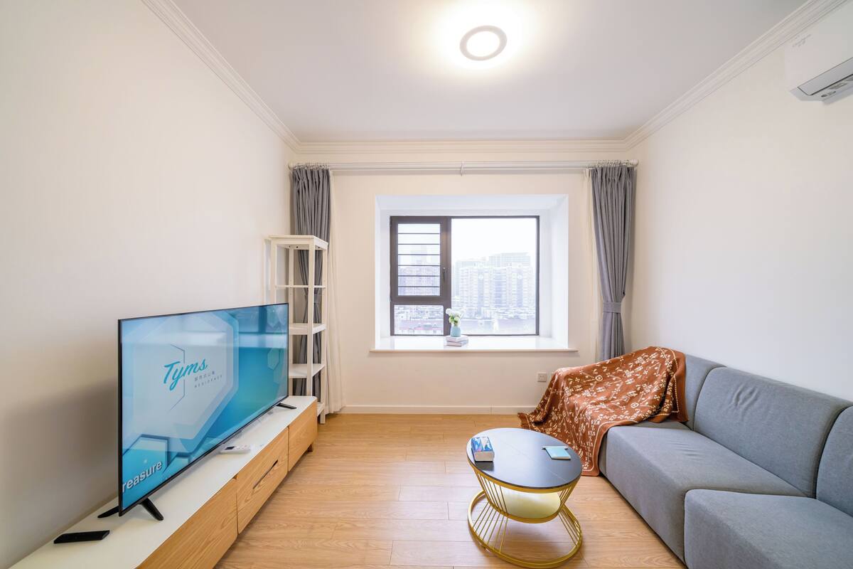 Uma sala de estar com televisão de tela plana, um sofá cinza, uma mesinha redonda e uma janela com cortinas.