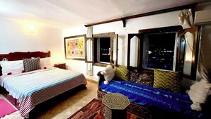 Quarto de hotel com duas camas, uma mesa de cabeceira, uma janela com vista para a cidade e um espelho decorativo.