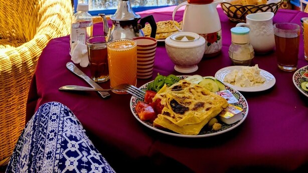 Uma mesa posta com comida e bebidas, uma cadeira de vime e vista para montanhas.