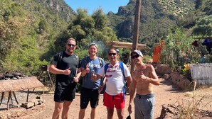 Quatro pessoas ao ar livre, com chão de terra, uma estrutura de madeira e uma montanha ao fundo.