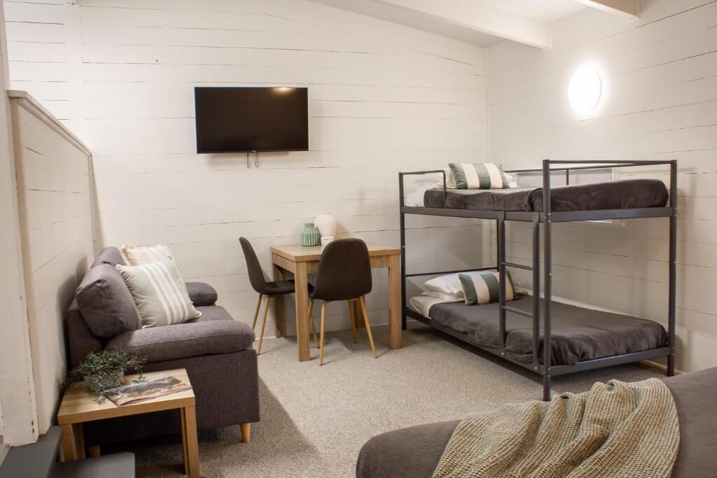 Quarto compacto com beliche, sofá, escrivaninha e televisão fixada na parede.