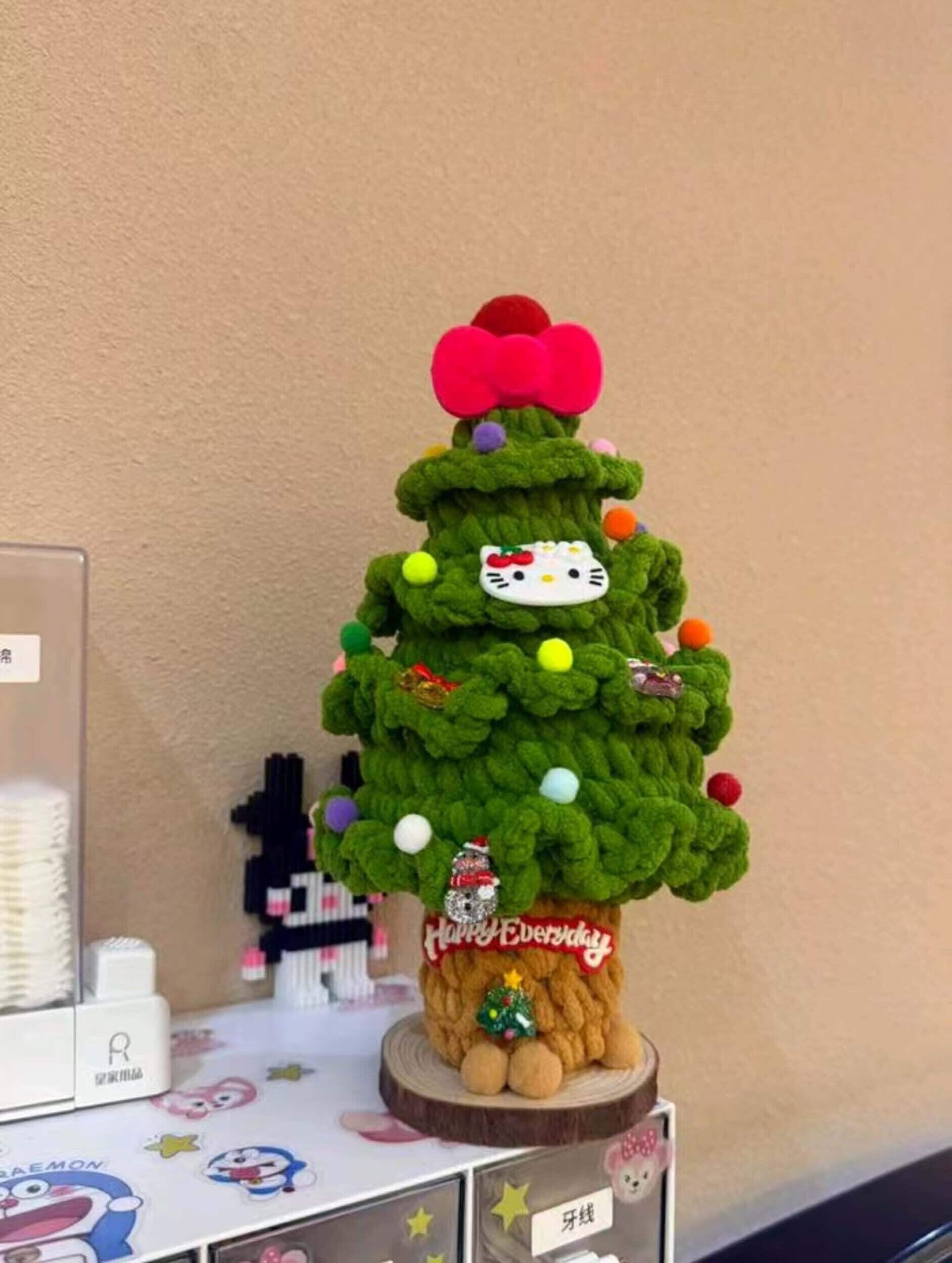 Uma árvore de Natal fofa do Hello Kitty com um laço vermelho em cima, colocada sobre um suporte de madeira.