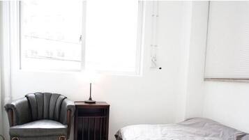 Quarto com uma cama de solteiro, uma cadeira, uma mesinha com abajur e uma janela com persianas.
