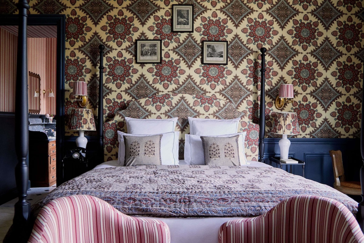 Quarto com papel de parede estampado, cama com almofadas, mesa de cabeceira, luminária e uma cadeira.