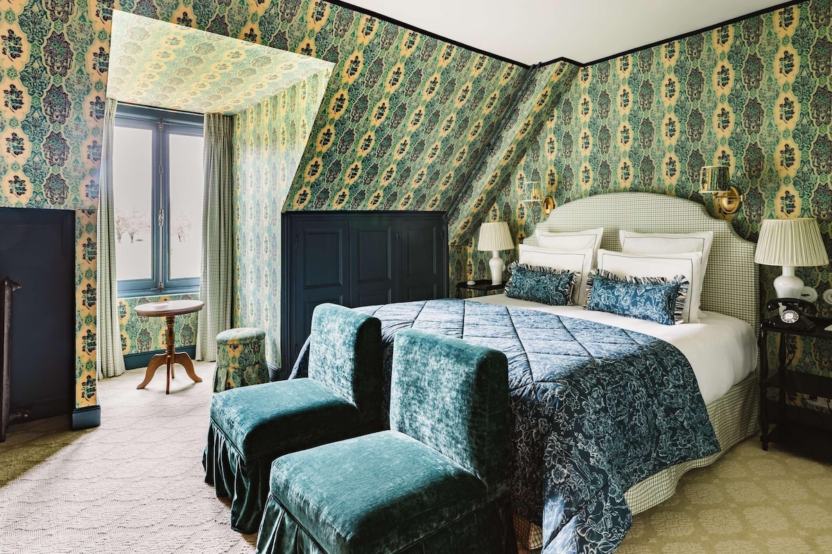 Quarto com cabeceira xadrez, cama com colcha estampada de flores azuis, duas poltronas combinando e uma mesinha redonda.