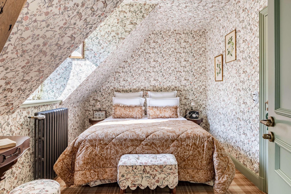 Um quarto com teto inclinado, papel de parede floral, uma cama com roupa de cama combinando, um radiador, um rádio antigo e uma pequena mesa.