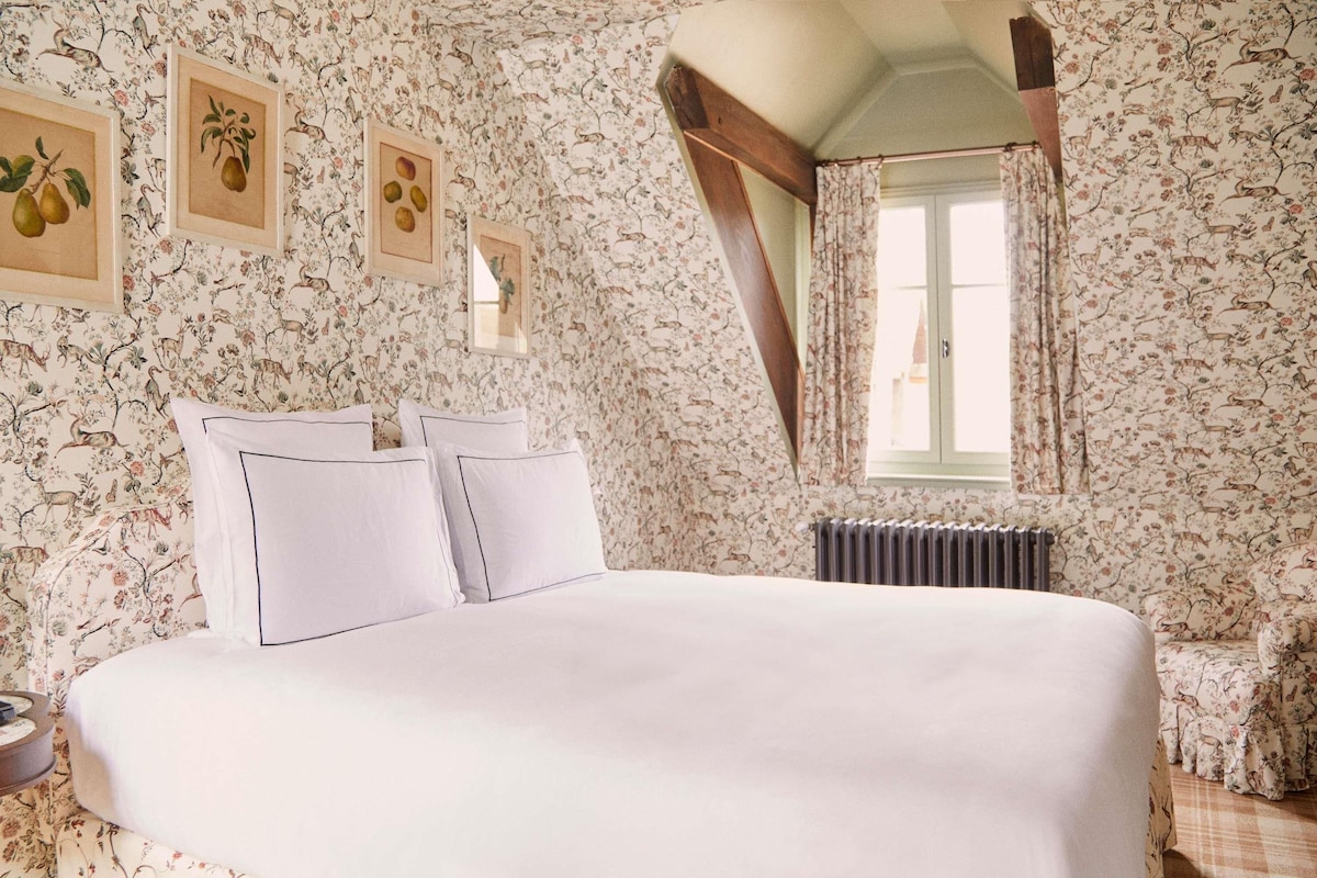 Quarto com cama grande, papel de parede floral, radiador e janela com cortinas.