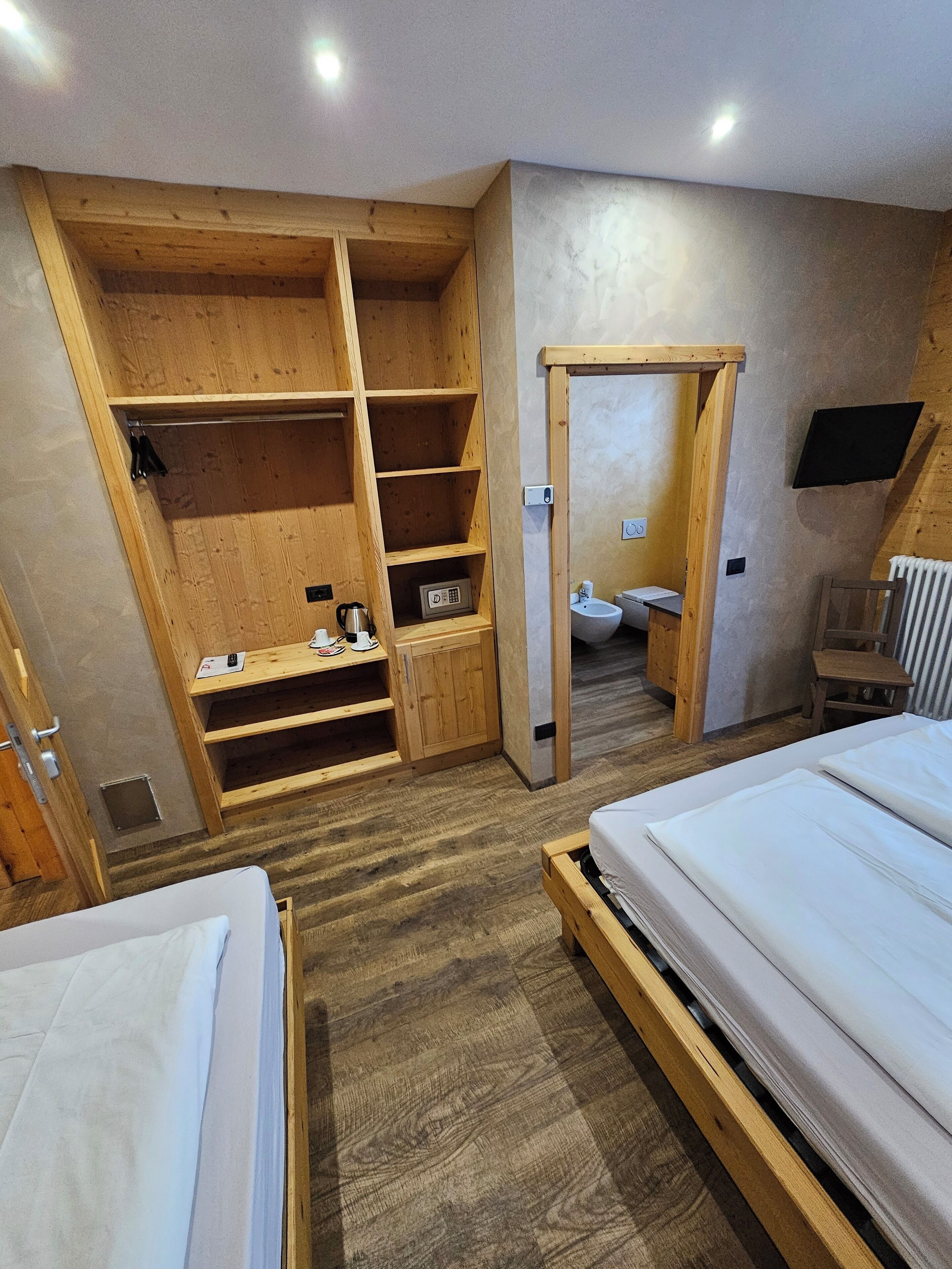 Quarto compacto com duas camas, guarda-roupa de madeira, banheiro pequeno e televisão na parede.