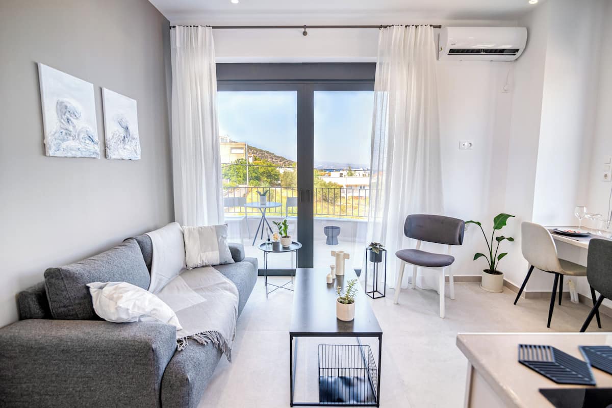 Uma sala de estar moderna com um sofá cinzento, uma mesa de centro, uma área de refeições com cadeiras e vista para a cidade através de grandes janelas.