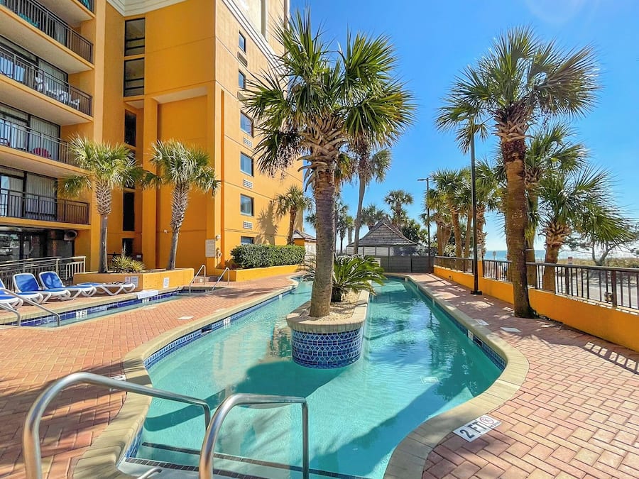 Uma área de piscina de hotel com piscina azul cristalina, espreguiçadeiras, palmeiras e fachada de um edifício amarelo.