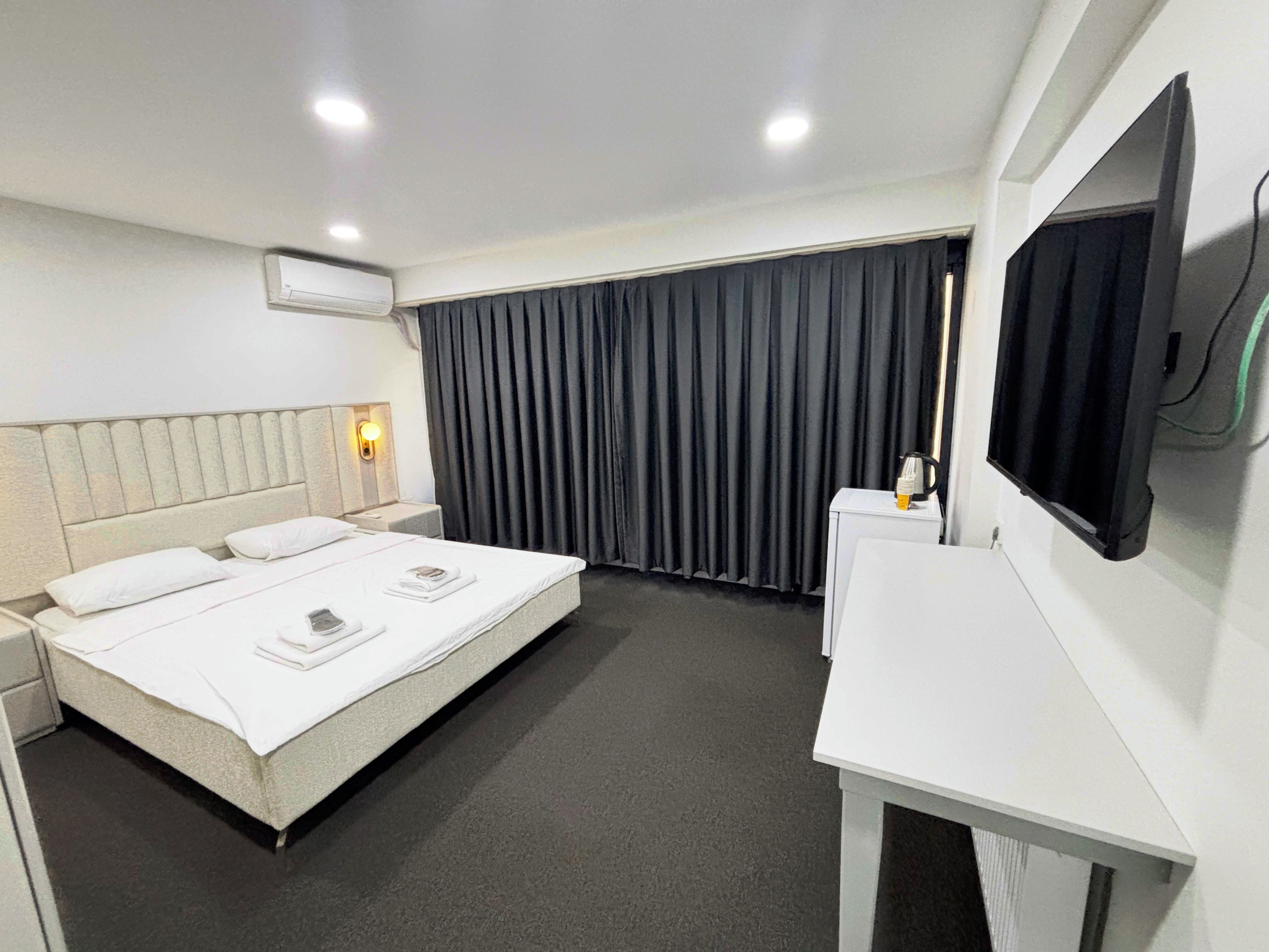 Quarto de hotel com cama, mesa de cabeceira, televisão e uma grande janela com cortinas.
