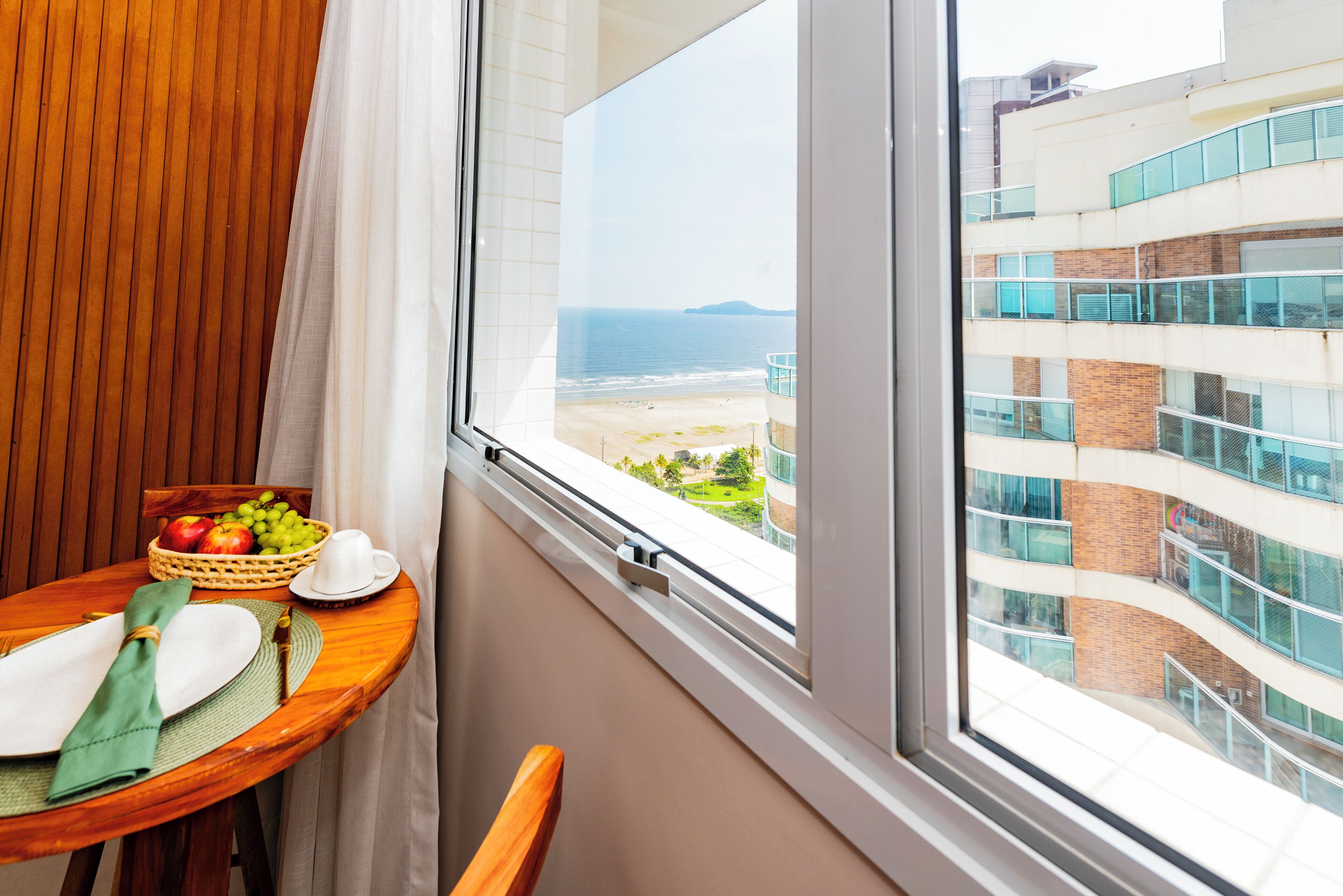 Quarto com vista para a praia e área de jantar com mesa posta para dois, cesta de frutas e uma janela com cortina.