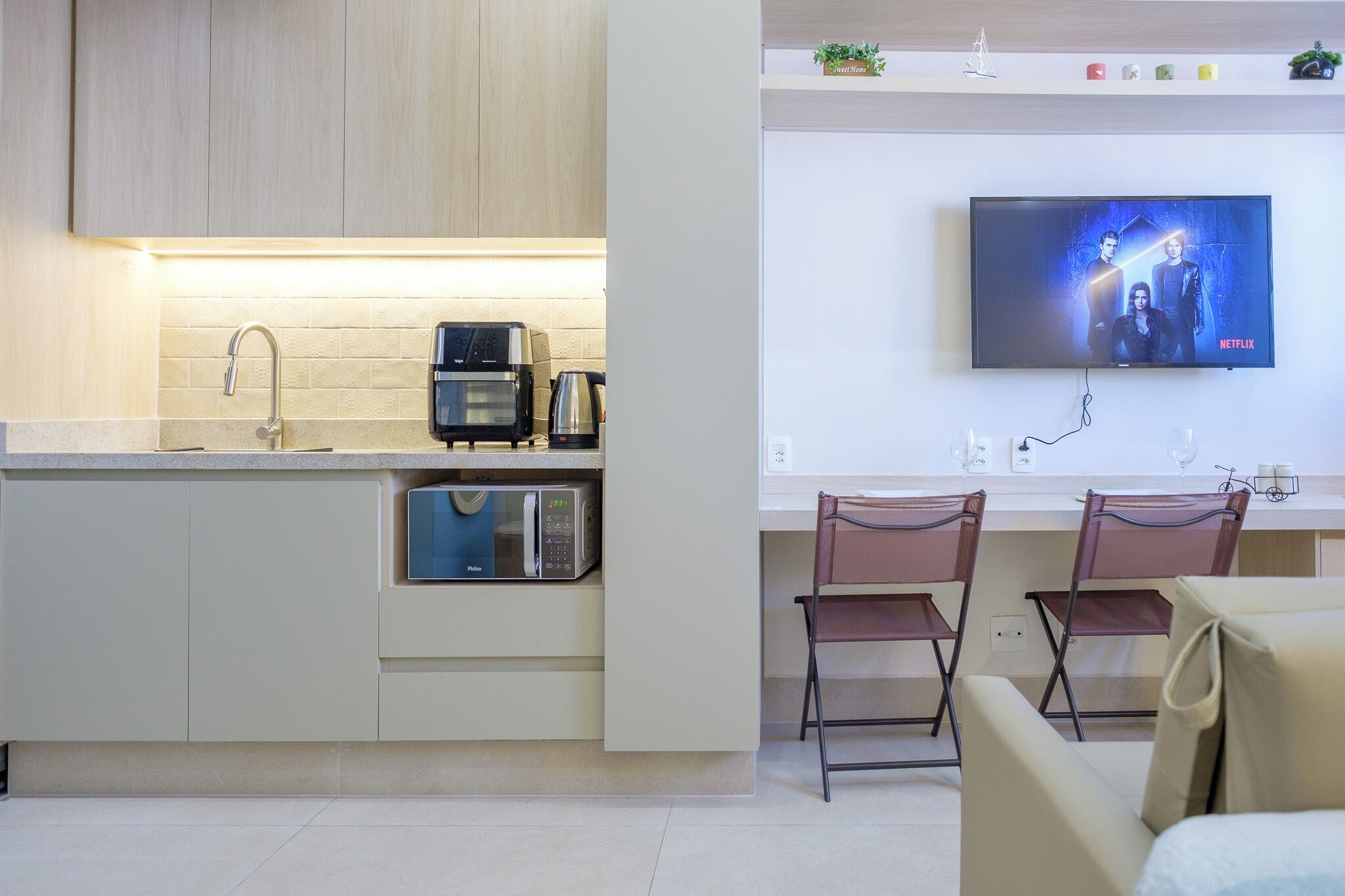 Cozinha moderna com micro-ondas, cafeteira e torradeira. Há uma TV na parede exibindo um programa. Duas cadeiras estão posicionadas à mesa de jantar.