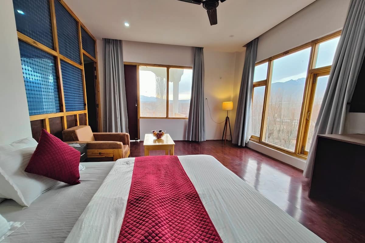 Quarto de hotel com uma cama grande, uma cadeira de madeira, uma mesinha com abajur e janelas amplas com vista para as montanhas.