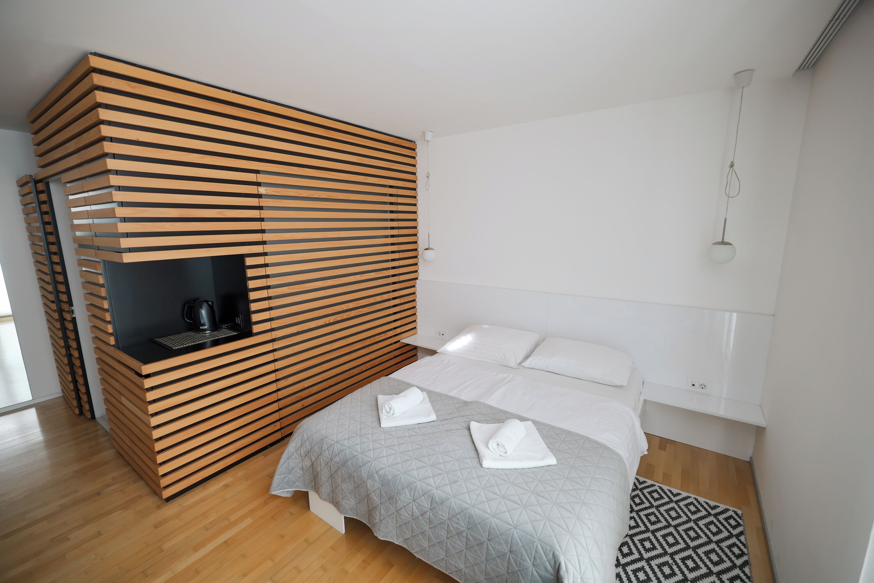 Um quarto moderno com parede de ripas de madeira, uma cama com roupa de cama branca e um tapete estampado.