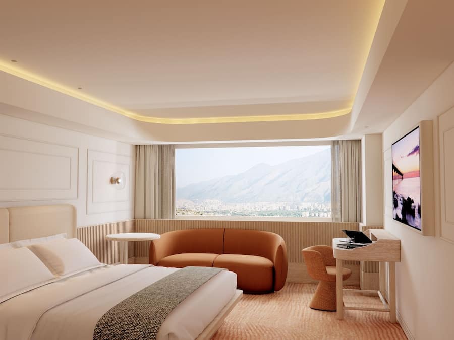 Quarto de hotel moderno com uma cama grande, um sofá, uma escrivaninha e vista para a paisagem urbana e montanhas.