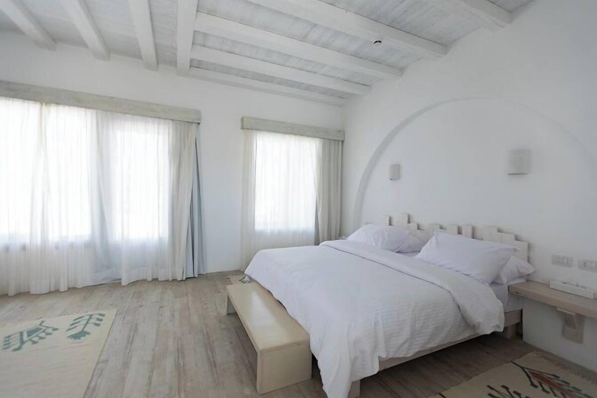 Quarto com cama, banco e luminária fixada na parede.