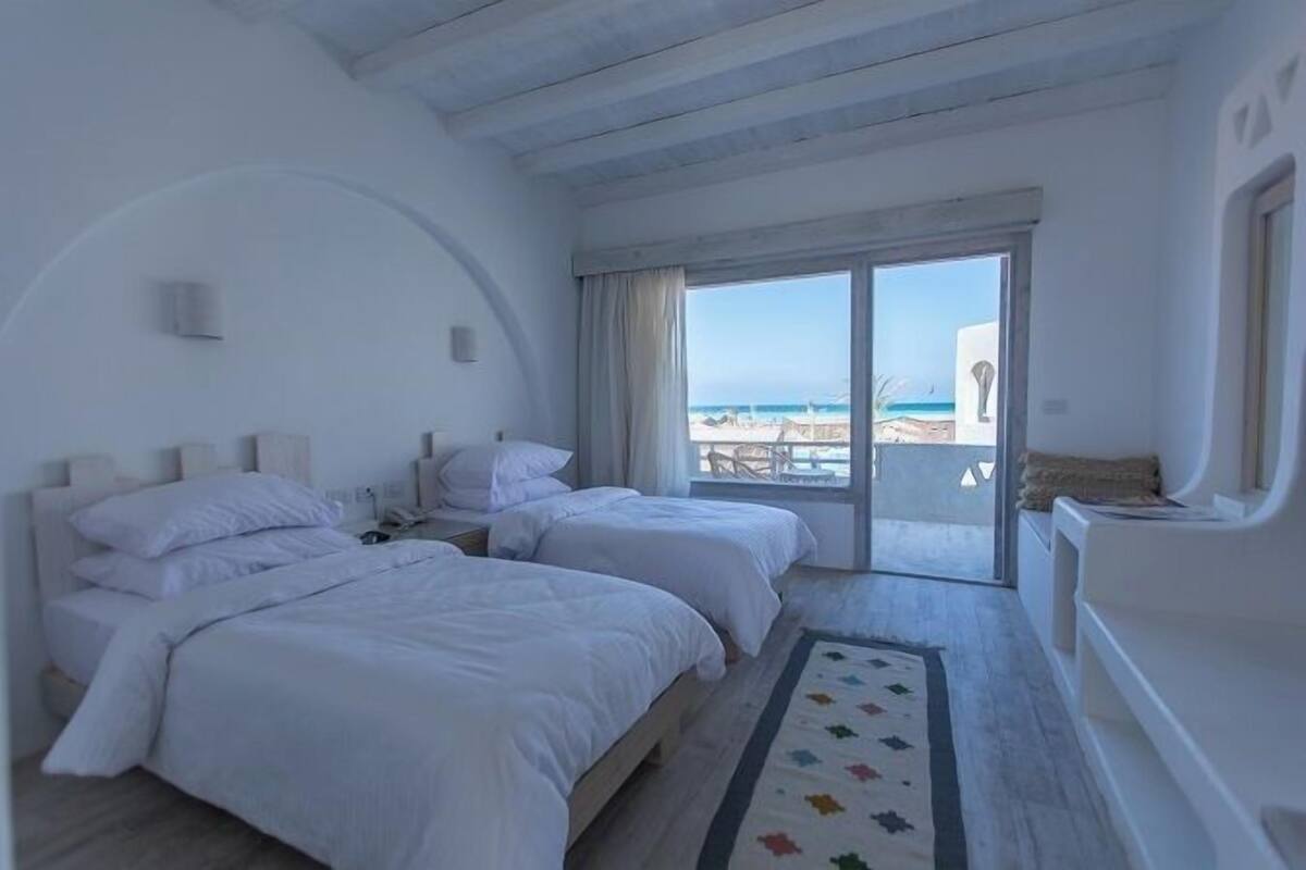 Quarto com duas camas, uma janela grande com vista para a praia e uma varanda com mesa e cadeiras.