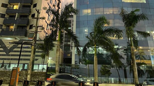 Um edifício moderno e imponente com fachada de vidro, rodeado por palmeiras e um carro estacionado.