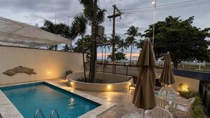 Piscina com uma escultura de mão, rodeada por um deck ladrilhado, com assentos externos e vista para o mar.